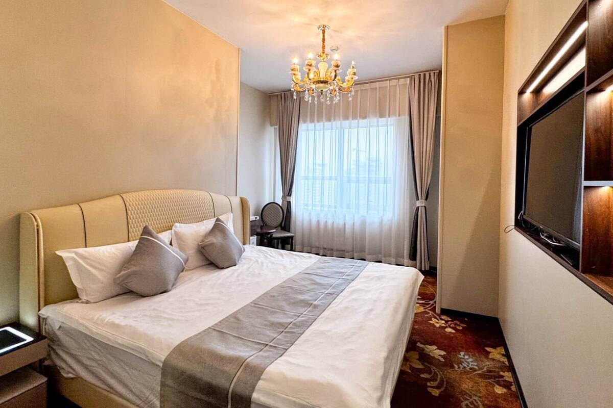 Quarto de hotel com cama grande, lustre, televisão e janela com cortinas.