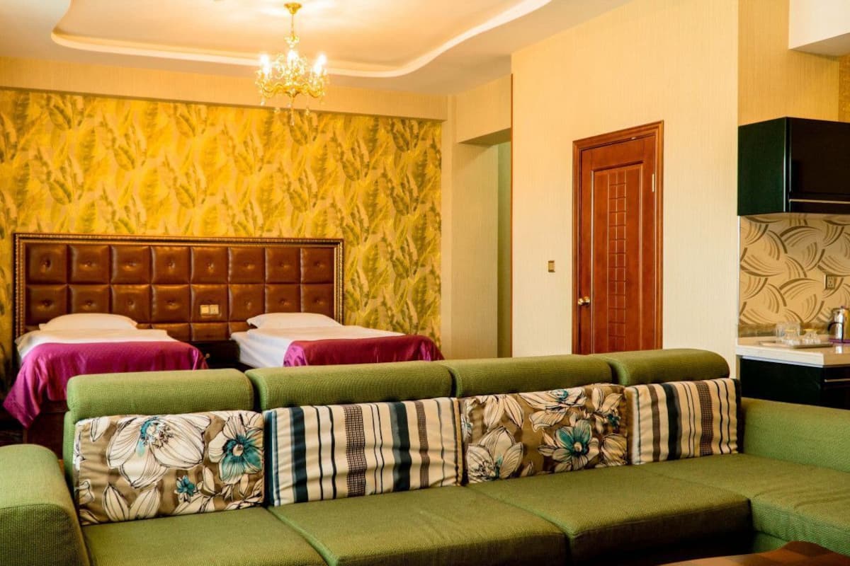 Quarto de hotel com um sofá verde, duas camas com roupa de cama vermelha e branca, uma porta de madeira e um candelabro.