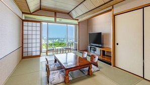 Um quarto em estilo japonês tradicional com tatames, uma mesa baixa com almofadas, uma televisão sobre suporte de madeira e uma porta deslizante que dá para uma varanda com vista para a cidade.