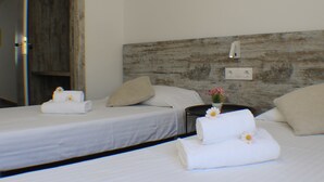 Quarto de hotel com duas camas, cada uma com colcha e almofadas brancas. Há uma pequena mesa com um vaso de flores e uma toalha.