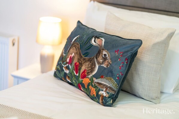 Uma cama com travesseiro com coelho e estampa floral, uma luminária de cabeceira e outro travesseiro texturizado na cama.