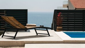 Uma área à beira da piscina com espreguiçadeira e uma mesa pequena, uma planta em vaso e vista para o mar.