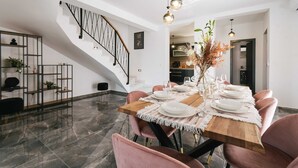 Uma área de refeições com uma mesa de madeira posta para seis pessoas, cercada por cadeiras cor-de-rosa, uma escada e uma cozinha moderna ao fundo.