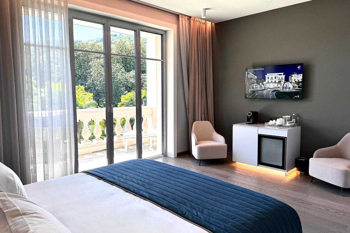Quarto de hotel com uma cama grande, uma escrivaninha com cafeteira, uma TV na parede e uma varanda com mobiliário de exterior.