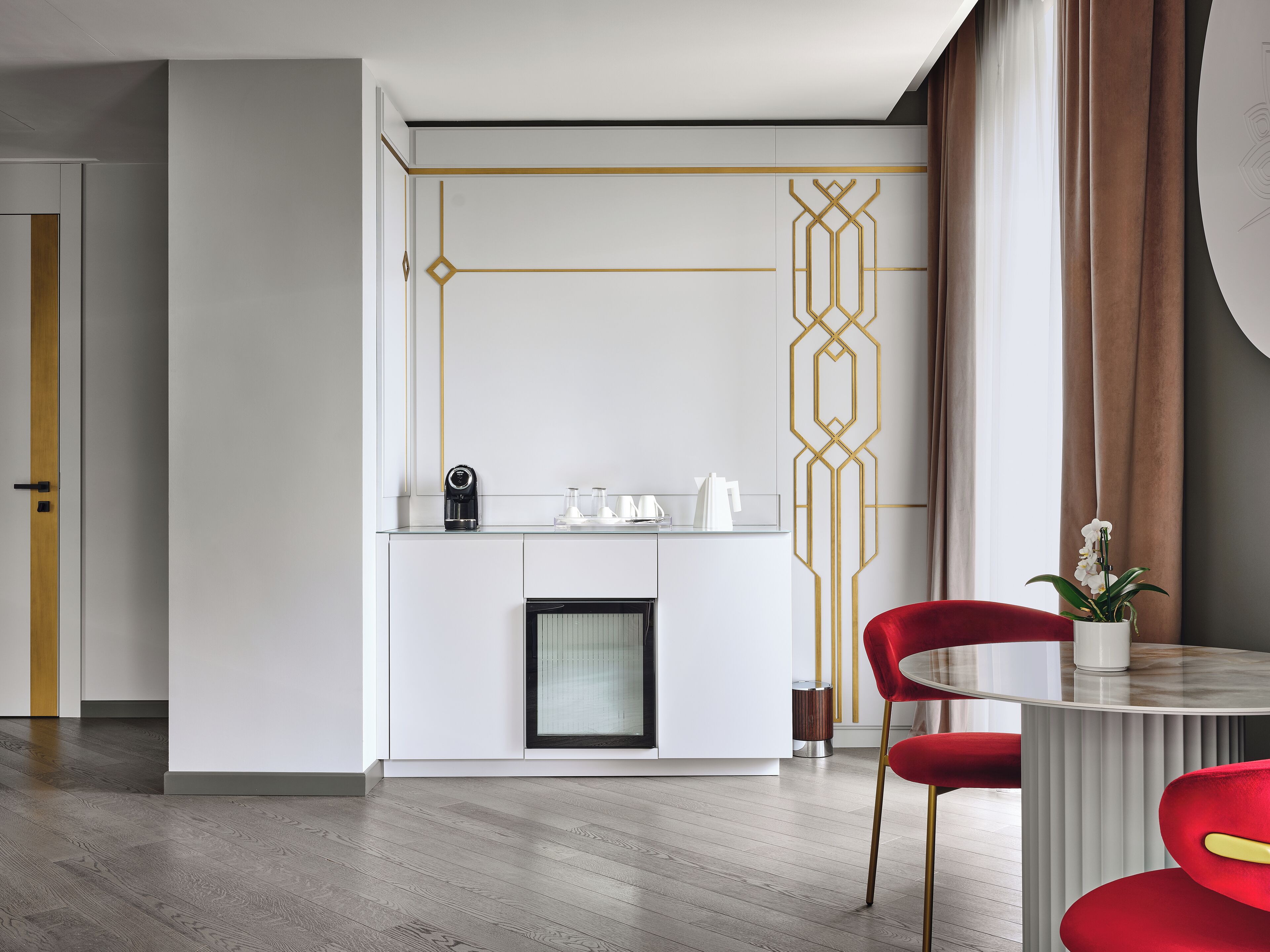 Quarto de hotel moderno com design minimalista, com uma pequena cozinha, área de jantar com cadeiras vermelhas e uma grande janela com cortinas.