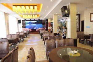 Uma área de refeições de hotel com várias mesas e cadeiras, uma tela grande exibindo uma vista panorâmica e um funcionário próximo à entrada.