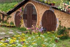 Uma casa no estilo Hobbit, com duas portas arredondadas, telhado de colmo e um jardim com diversas flores.