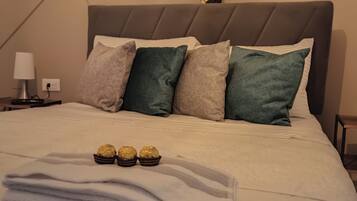Cama bem arrumada com uma toalha branca dobrada e três chocolates embalados, mesa de cabeceira com luminária e uma figura decorativa na cabeceira.