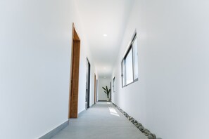 Um corredor comprido e estreito, com paredes brancas e chão cinzento, com molduras de porta em madeira e grandes janelas de um lado.