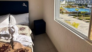 Quarto de hotel com uma cama, uma janela com vista para uma piscina e outros edifícios, e um pequeno objeto preto no chão.