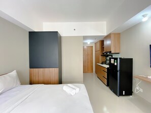 Quarto de hotel com cama, guarda-roupa, cozinha compacta com frigorífico e porta de madeira que dá para outro espaço.