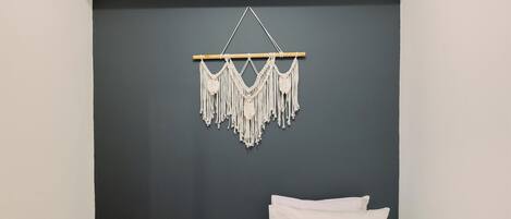 Cama com roupa de cama e travesseiros brancos, em frente a uma parede escura, com um painel de macramé pendurado acima.