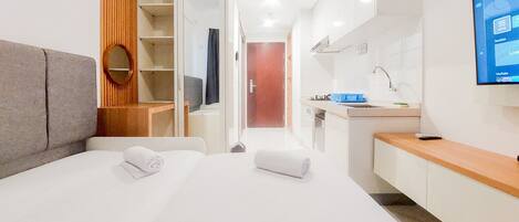 Quarto de hotel moderno com uma cama, um sofá cinza, uma estante de madeira, uma televisão e um banheiro com pia e espelho.