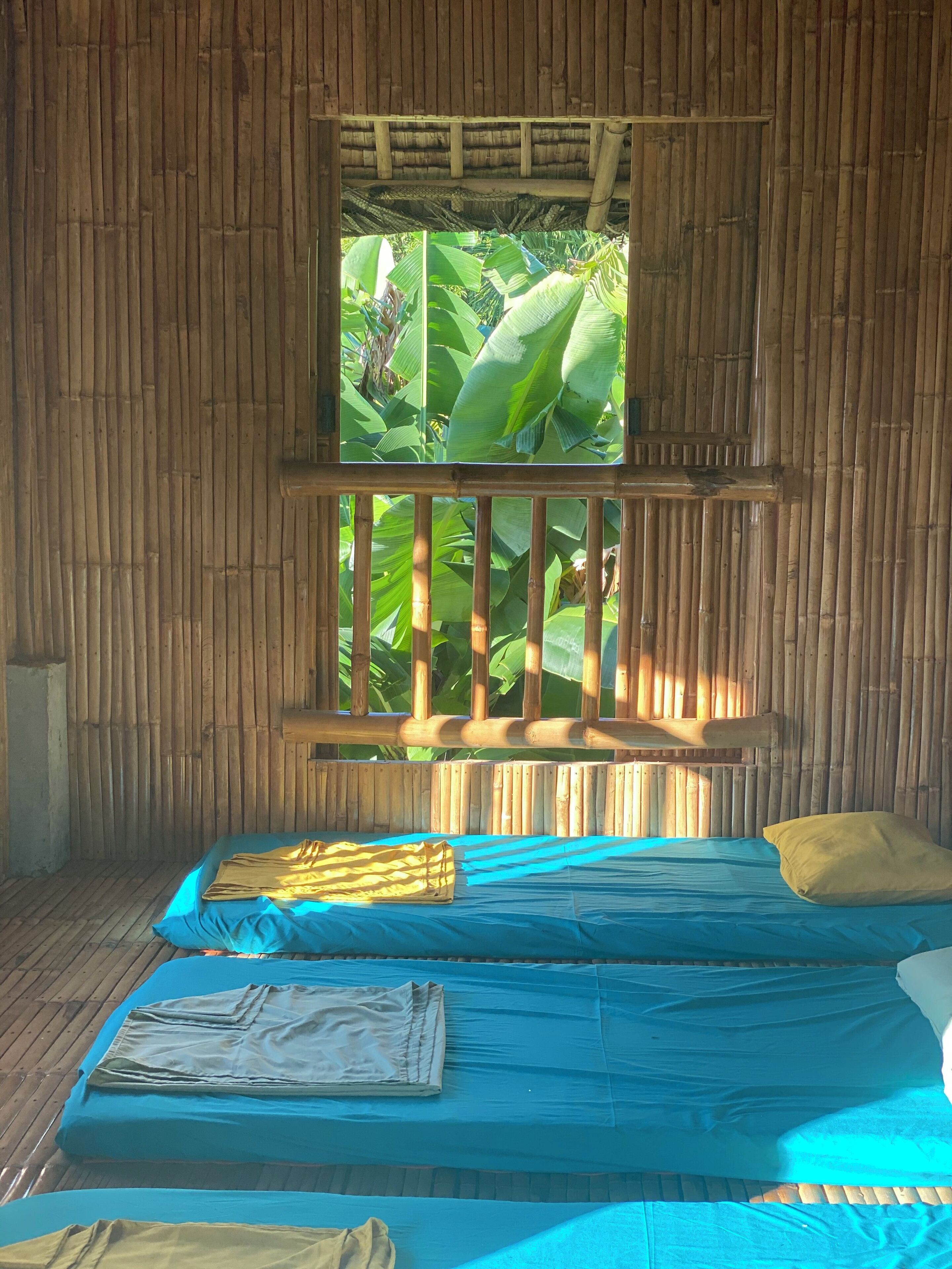 Quarto com parede de bambu, janela com vista para folhagem verde, cama com lençóis azuis e um banco de madeira.