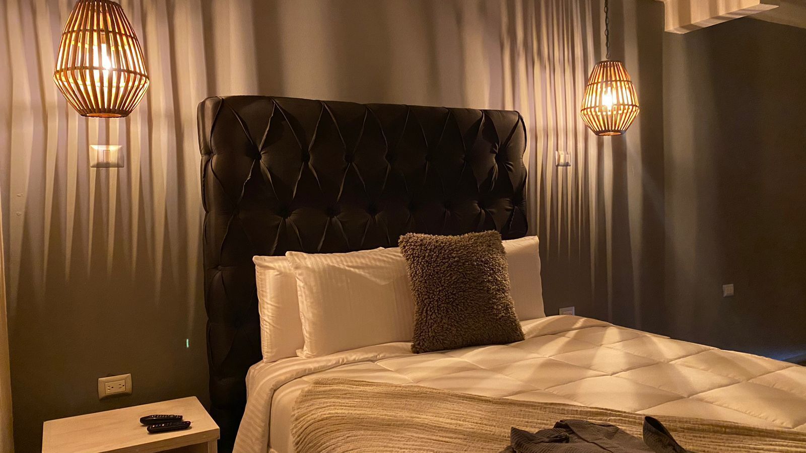 Quarto de hotel com cabeceira estofada, cama com roupa de cama branca, duas luminárias fixadas na parede e mesa de cabeceira com um controle remoto.