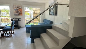 Um interior moderno com uma escada que leva a um segundo andar, uma sala de jantar com mesa e cadeiras, e uma sala de estar com sofá e um quadro na parede.