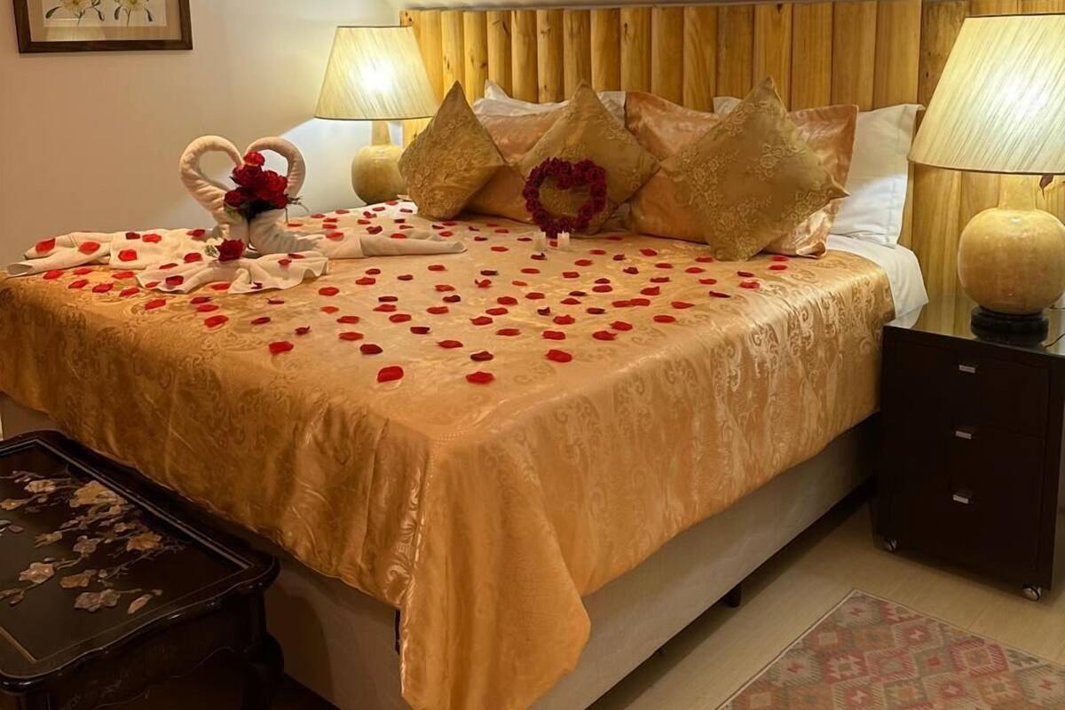 Cama bem arrumada com pétalas de rosa e um arranjo em formato de coração, cabeceira de madeira, mesa de cabeceira com abajur e um quadro na parede.