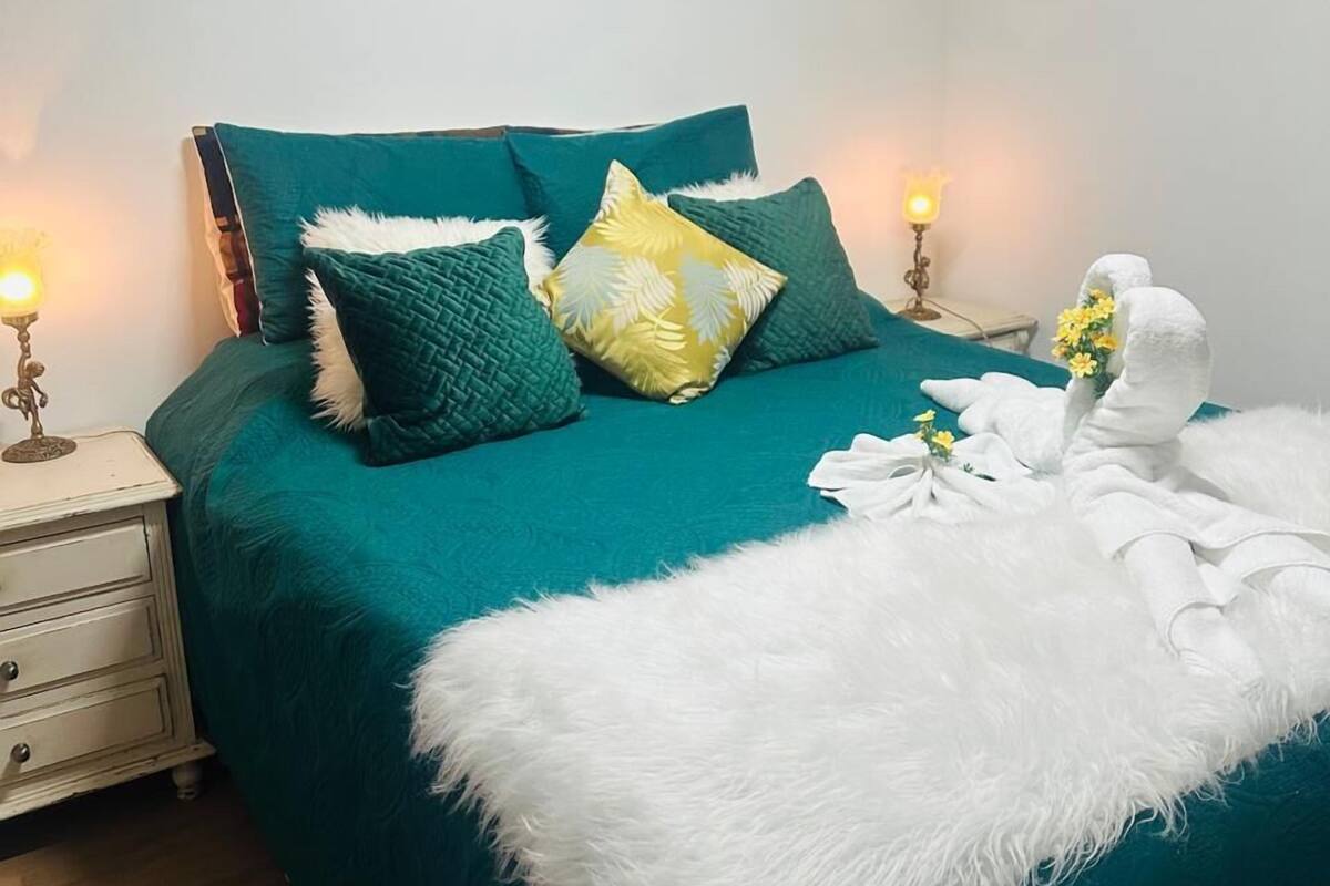 Cama bem arrumada com roupa de cama verde-água e almofadas brancas e fofas, um cobertor de penas branco e uma mesa de cabeceira com uma luminária.
