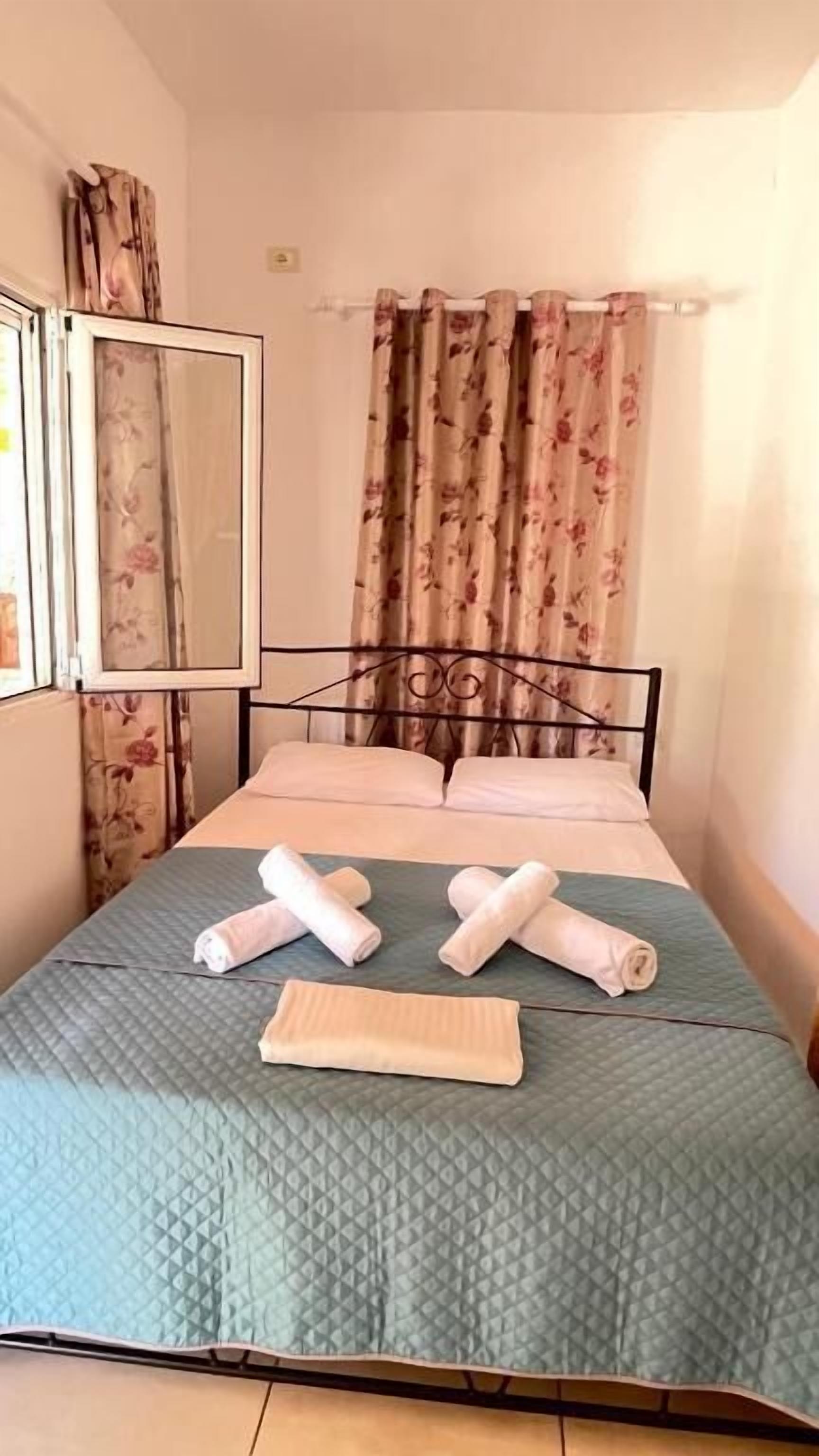 Uma cama de solteiro com colcha verde acolchoada, duas toalhas enroladas e cabeceira de metal.