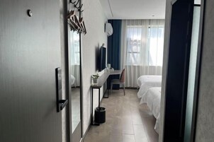Quarto de hotel com uma cama, uma escrivaninha, uma cadeira, uma mesinha com uma planta, um espelho e uma janela com cortinas.