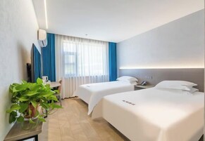 Quarto de hotel com duas camas, uma escrivaninha, uma cadeira, uma planta em vaso e uma janela com cortinas.