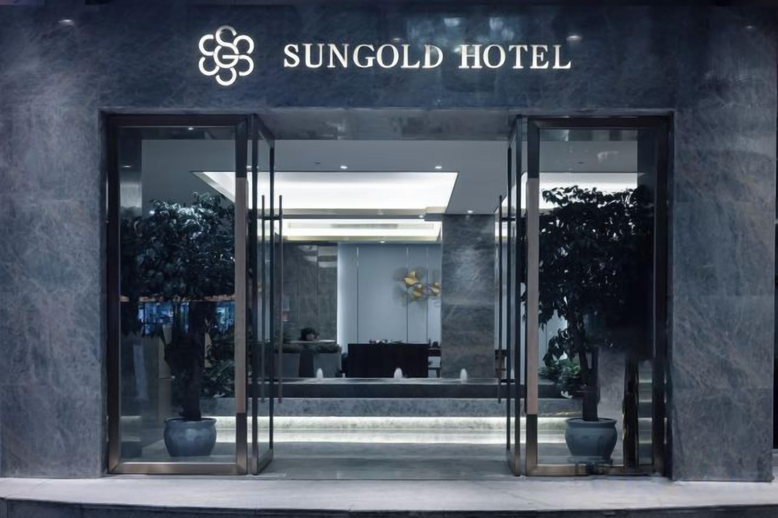 Entrada do Hotel Sungold, com porta de vidro, balcão de recepção no interior e plantas em vasos do lado de fora.