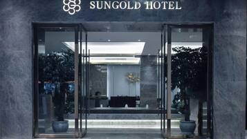 Entrada do Hotel Sungold, com porta de vidro, balcão de recepção no interior e plantas em vasos do lado de fora.