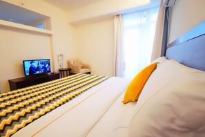 Quarto de hotel com cama, televisão, cadeira e janela com cortinas.