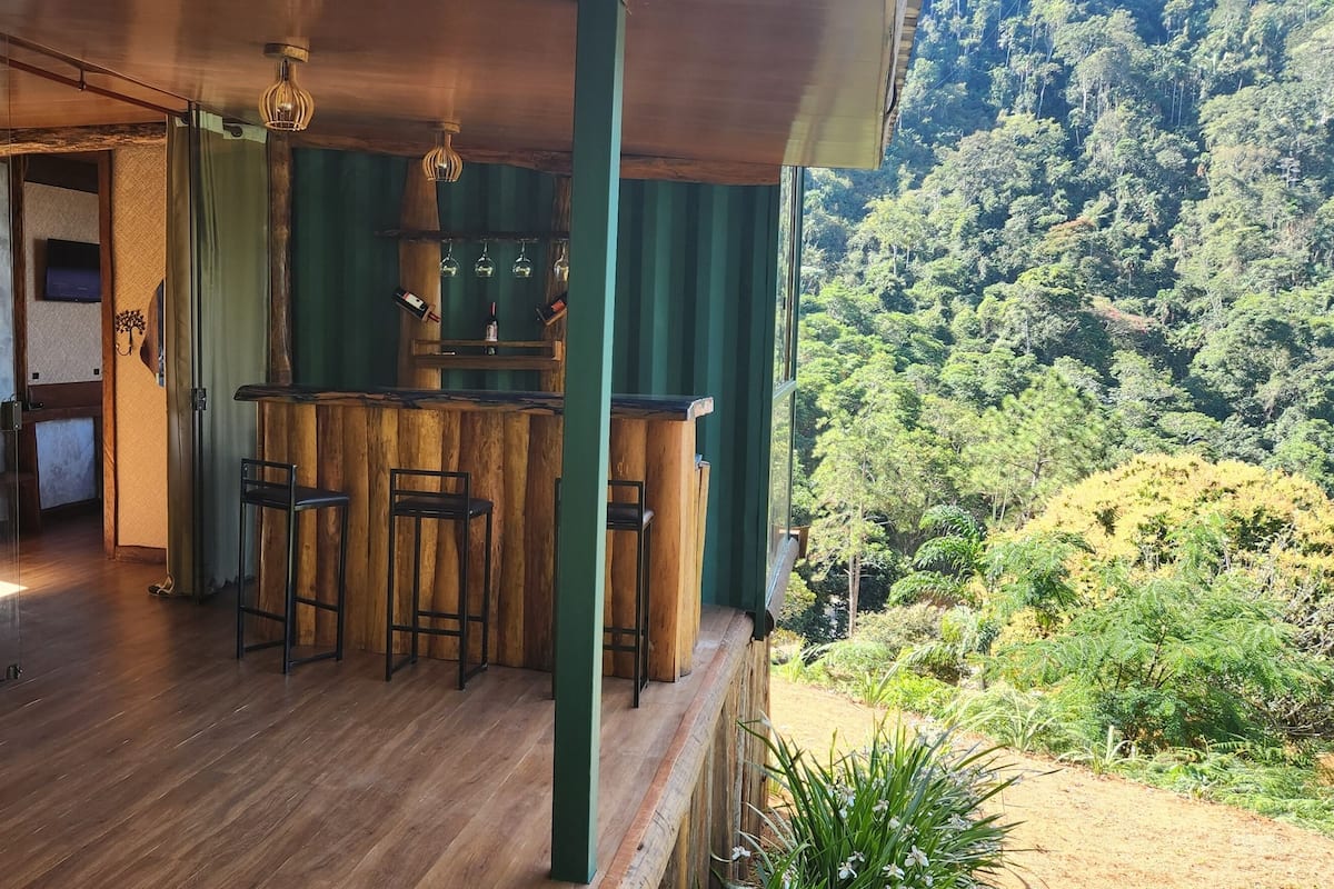 Um deck de madeira com área de bar, uma porta de vidro que dá para um espaço interno e vista para uma encosta arborizada.