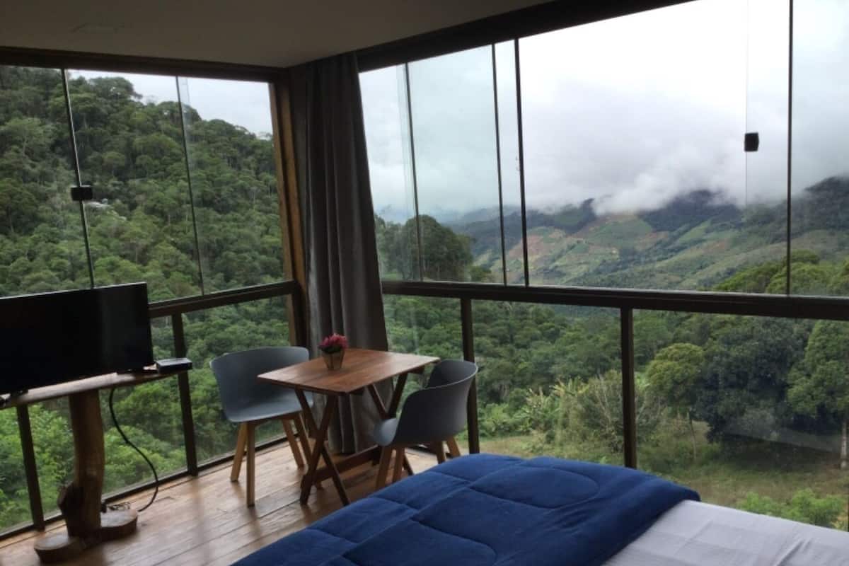 Quarto com uma janela grande, uma cama, uma televisão e uma pequena mesa com cadeiras.