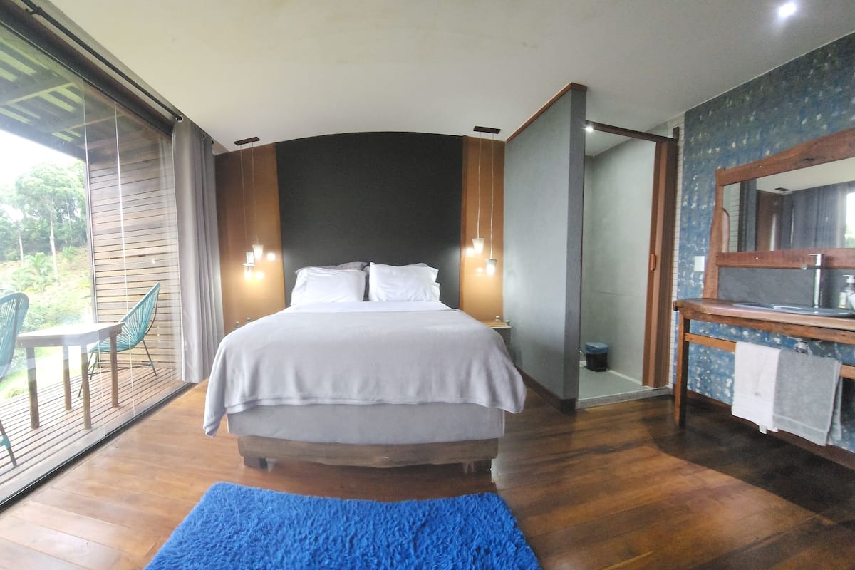 Quarto com cama, mesa de cabeceira, espelho, tapete azul e varanda com cadeiras.