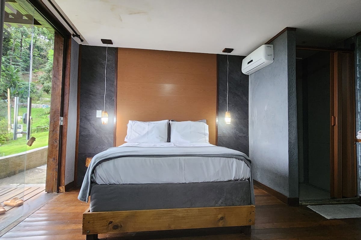 Quarto com cama de madeira, mesa de cabeceira, espelho e vista para o exterior.