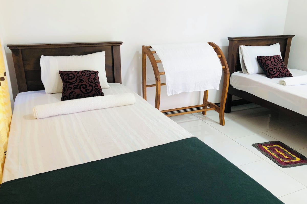 Quarto de hotel com duas camas individuais, cada uma com cabeceira de madeira, roupa de cama branca e almofadas estampadas. Uma cadeira de madeira com toalha branca está colocada entre as camas.