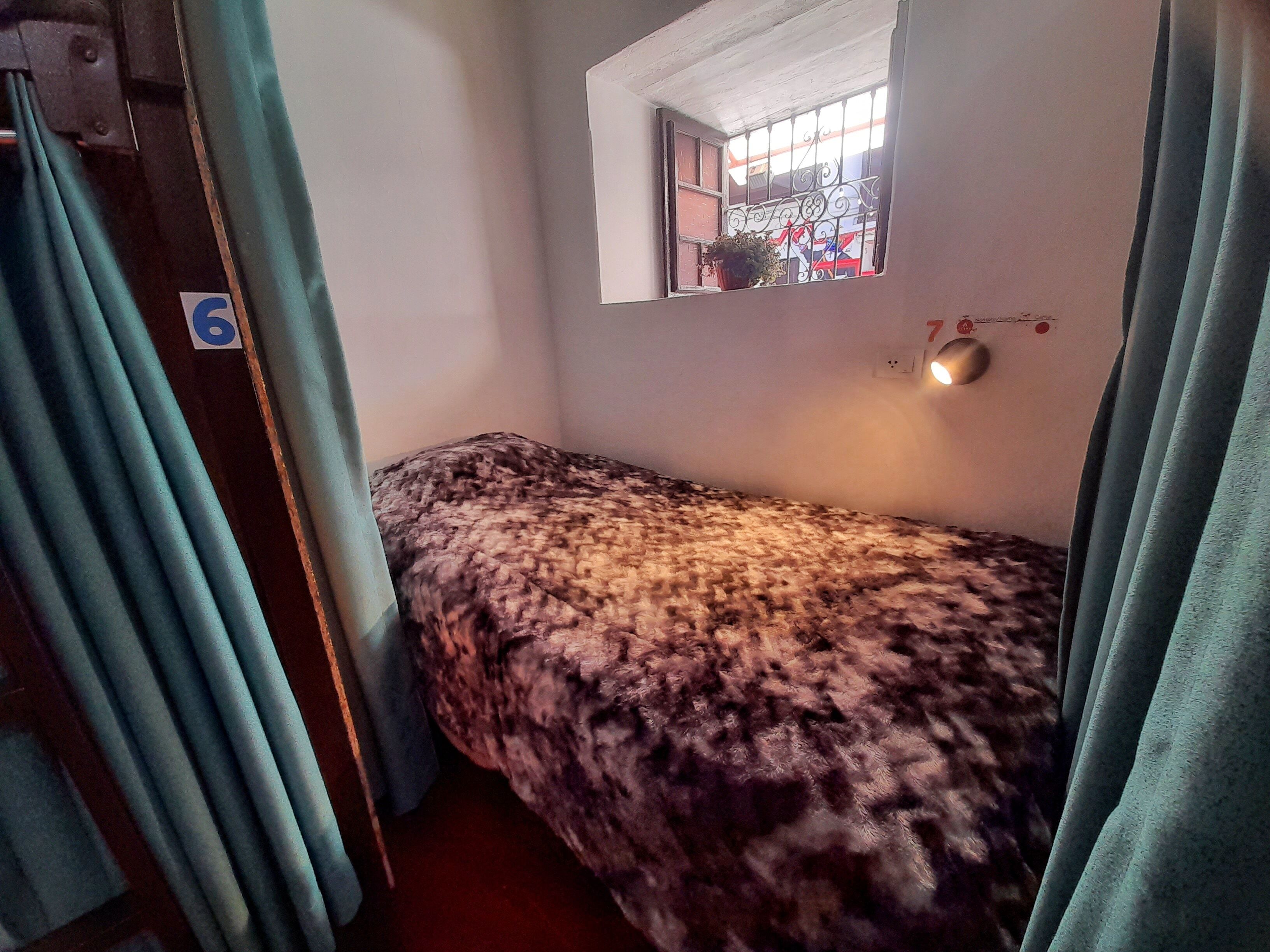 Um quarto pequeno e estreito, com uma cama de solteiro, uma janela com grade de metal e um ponto de luz fixado na parede.