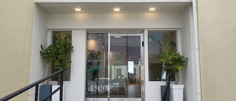 Entrada de um edifício com porta de vidro, escadas e plantas em vasos.