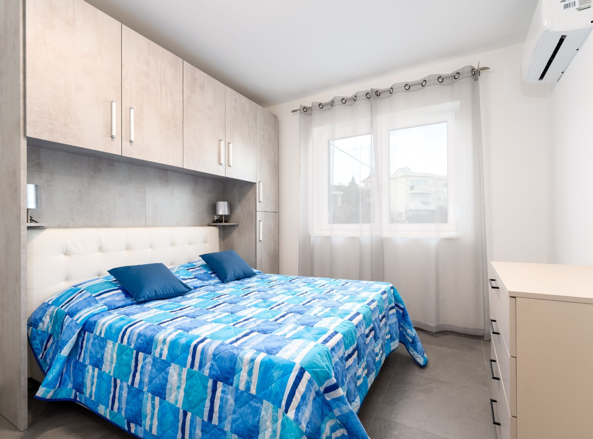 Quarto com cama, guarda-roupa, janela com cortinas e aparelho de ar condicionado.
