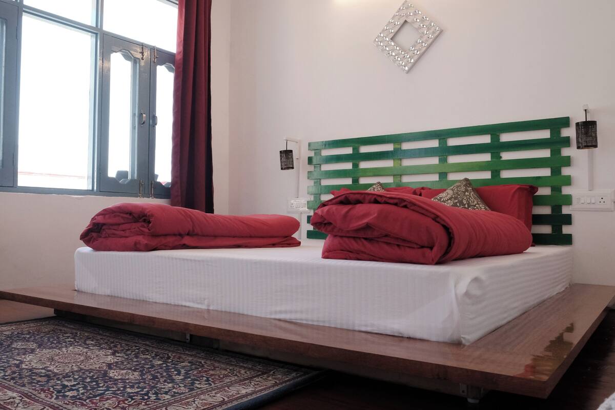 Uma cama com cabeceira verde, roupa de cama vermelha e um tapete estampado sobre uma plataforma de madeira.