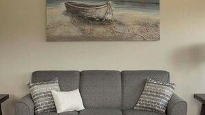 Um sofá cinzento com almofadas estampadas, uma mesa de centro e um quadro de um barco numa praia.