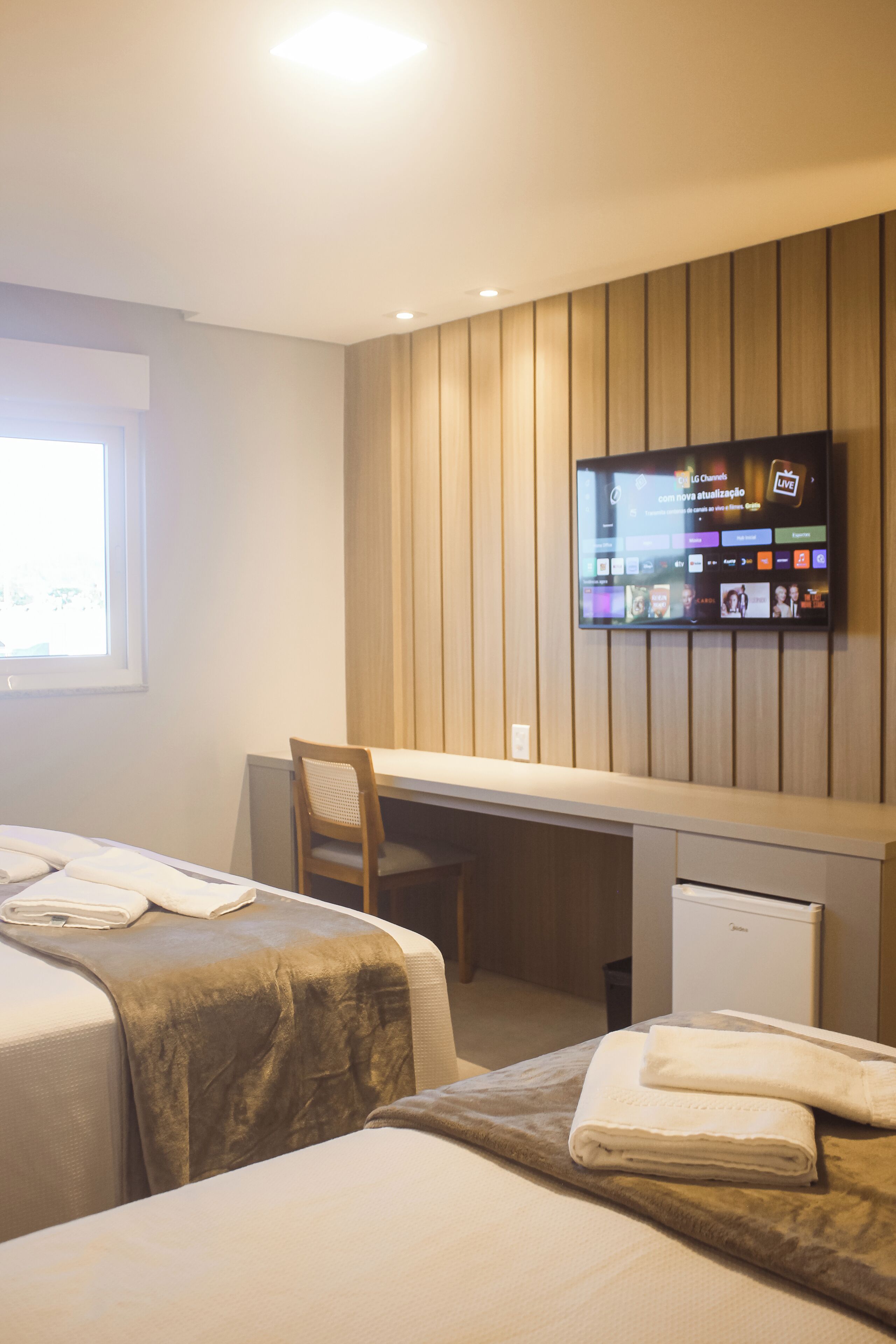 Quarto de hotel com cama, escrivaninha, cadeira e televisão fixada na parede.
