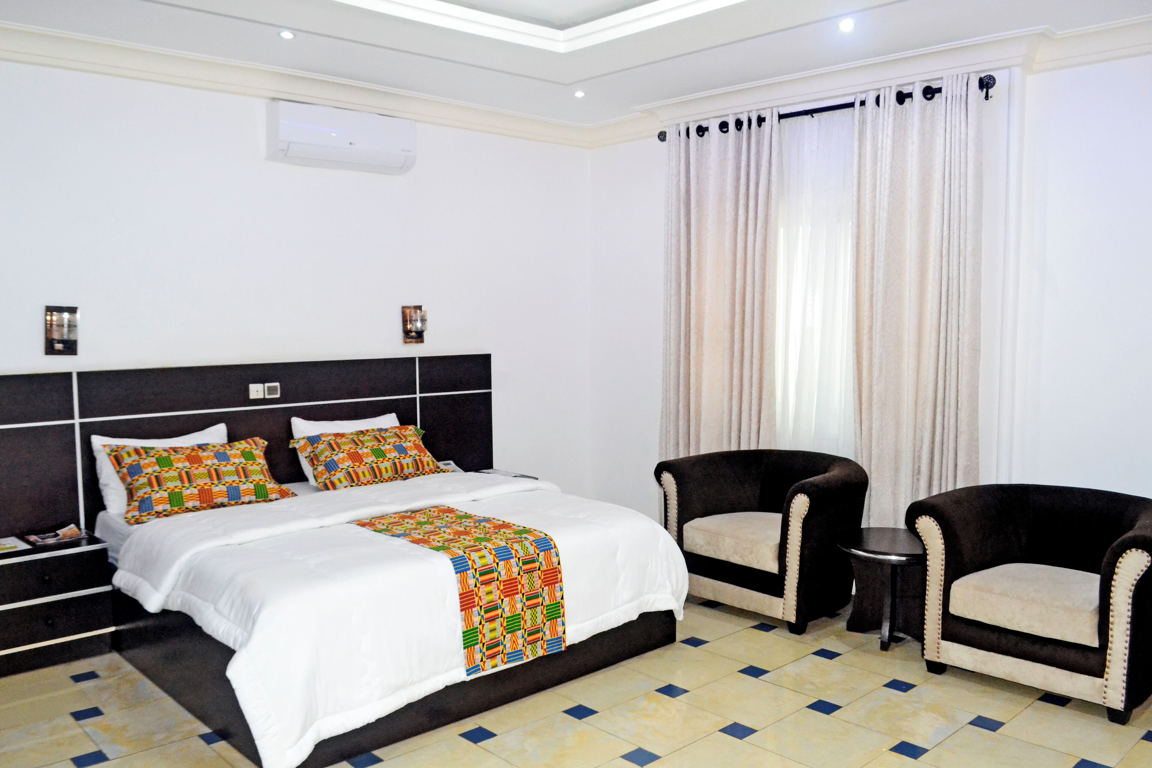 Quarto de hotel com uma cama, duas poltronas, uma mesa pequena, uma mesa de cabeceira e uma janela com cortinas.