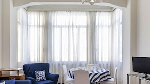 Quarto de hotel com uma grande janela, uma poltrona azul, uma mesa listrada, uma televisão e um quadro na parede.