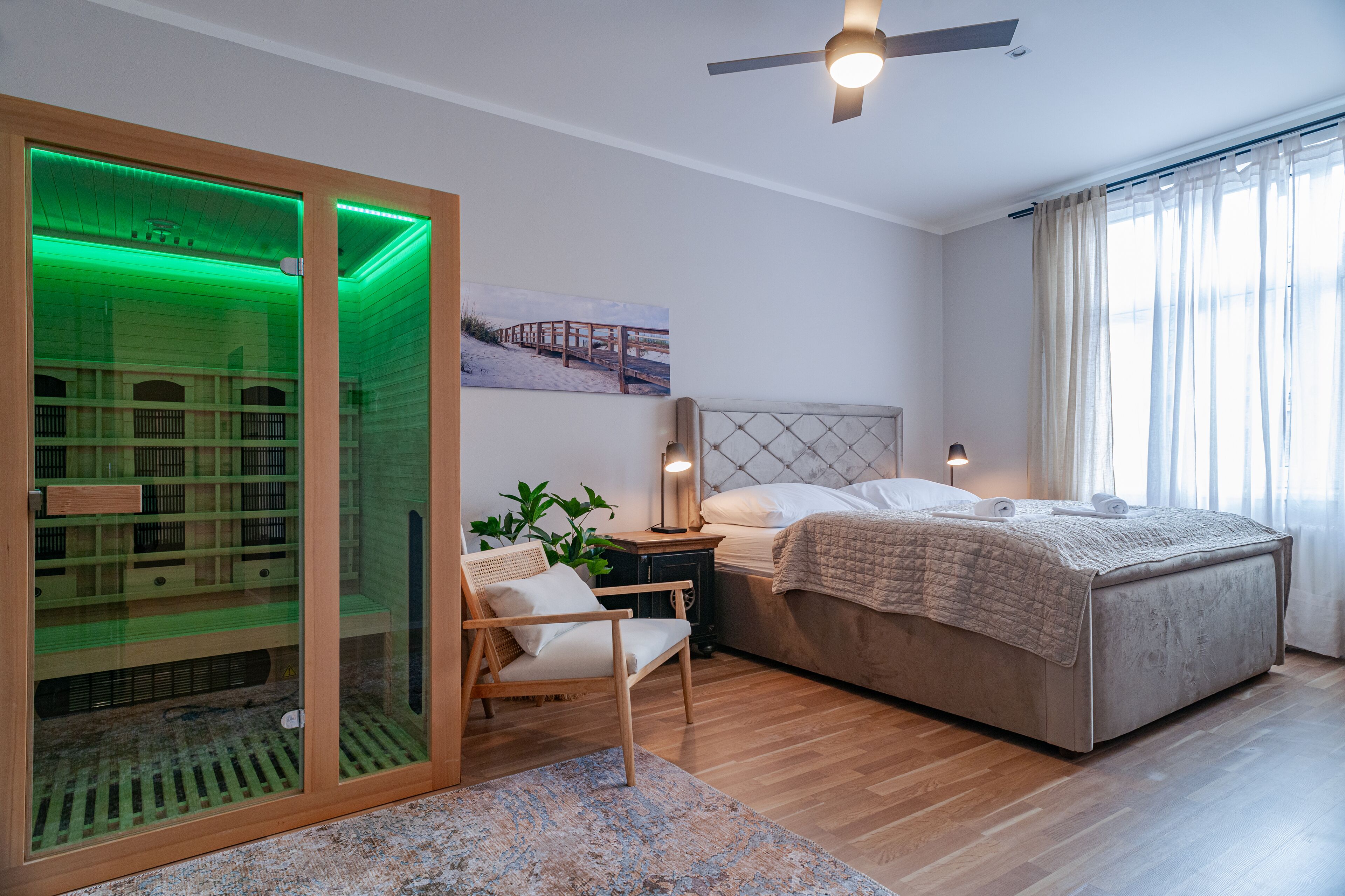 Quarto com cama, cadeira, mesa de cabeceira, luminária, pintura e janela com cortinas.
