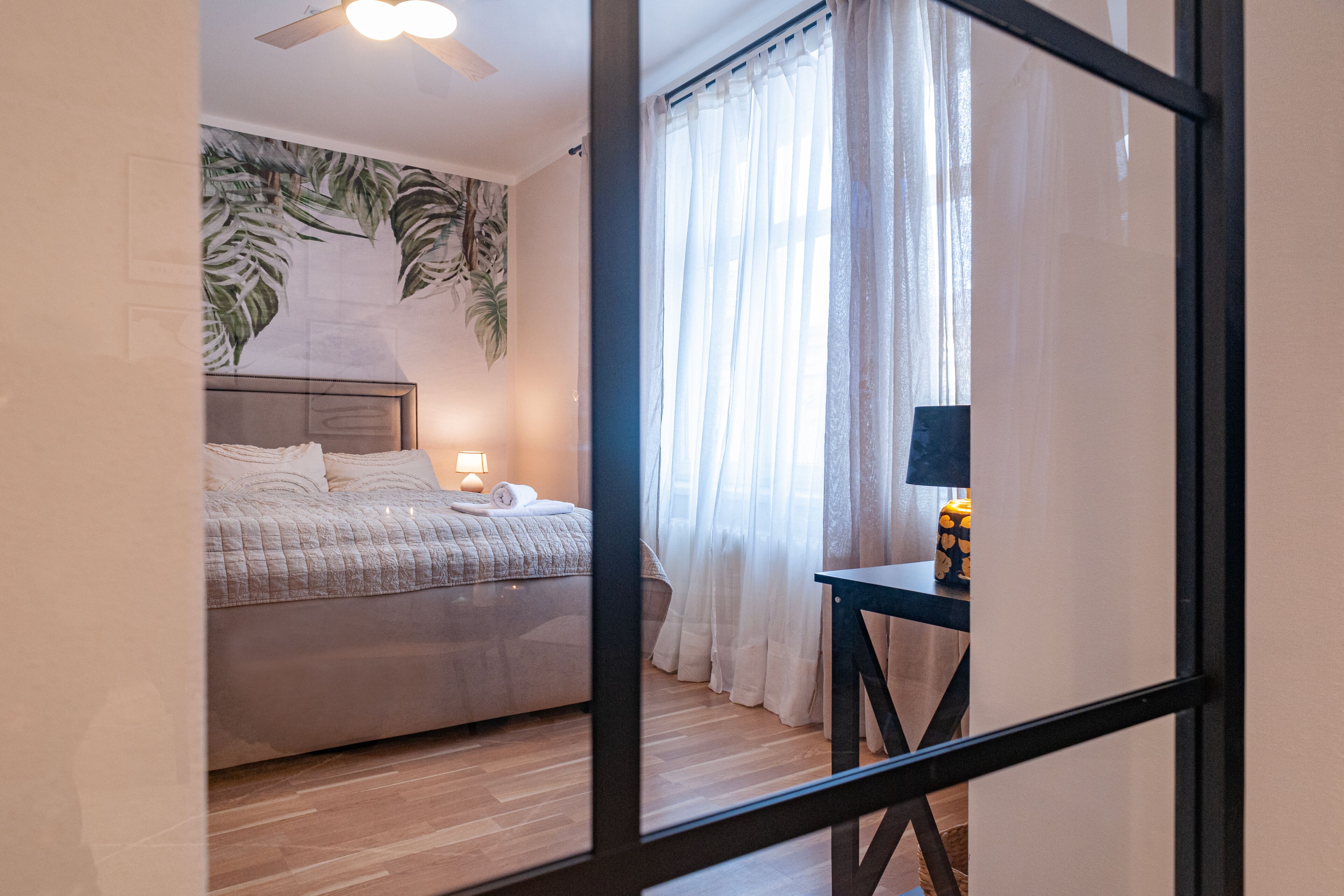 Quarto com cama, mesa de cabeceira, luminária e vista pela janela com cortinas.