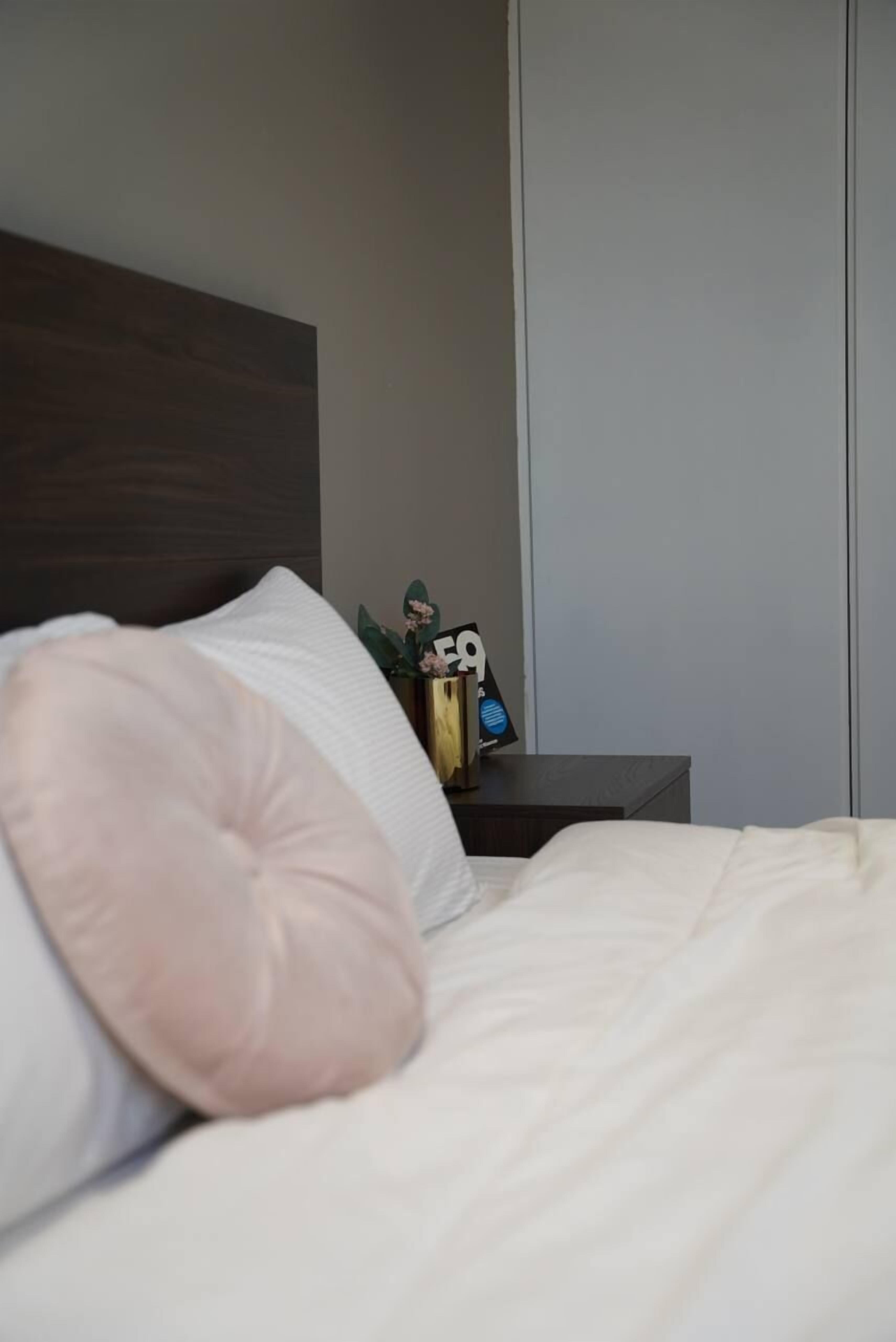 Cama bem arrumada com edredom branco e travesseiro rosa. Uma pequena mesa de cabeceira com um vaso dourado e um objeto decorativo é visível.