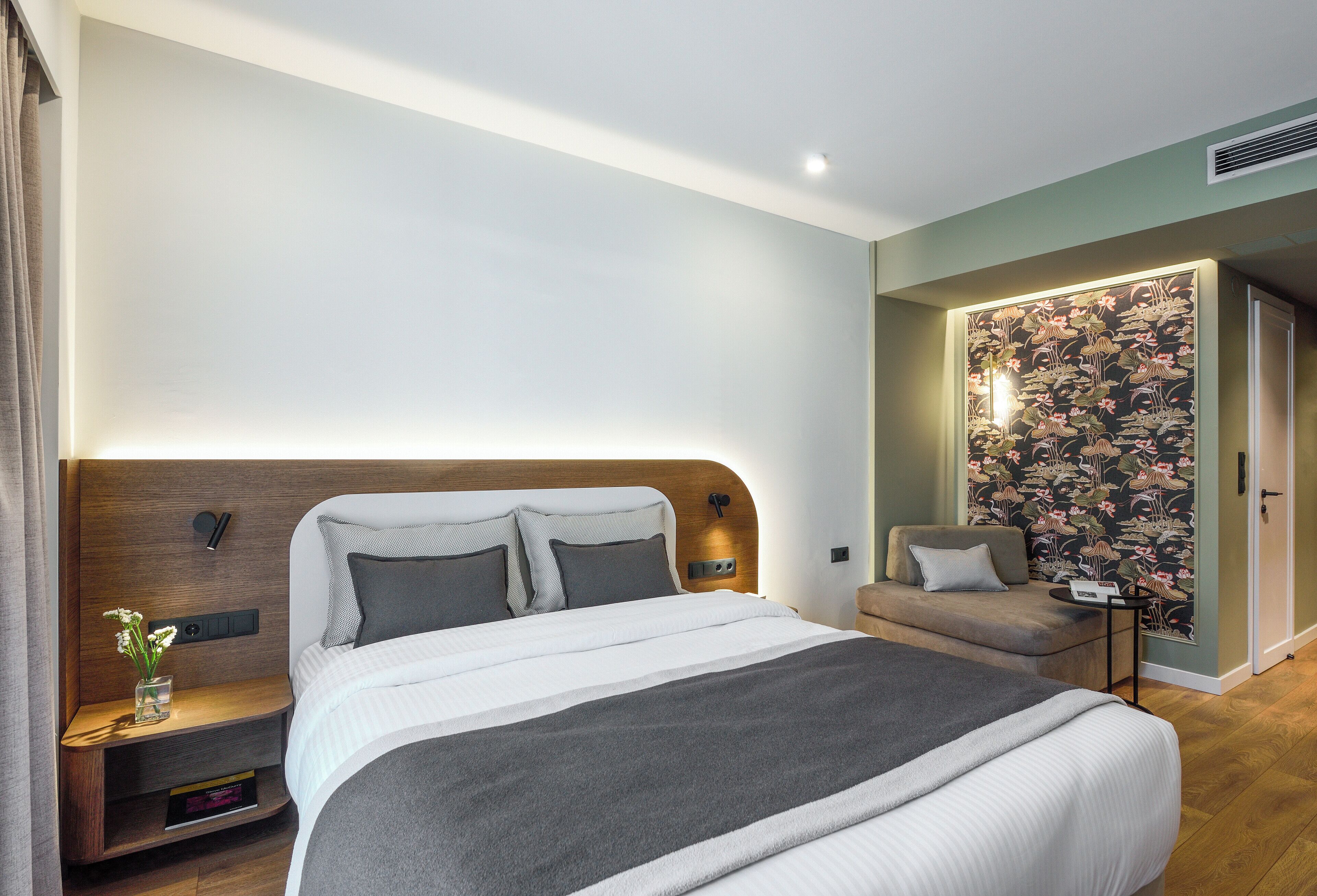 Quarto de hotel moderno com uma cama grande, mesas de cabeceira, um criado-mudo, uma pequena mesa com uma luminária e parede com papel de parede floral.