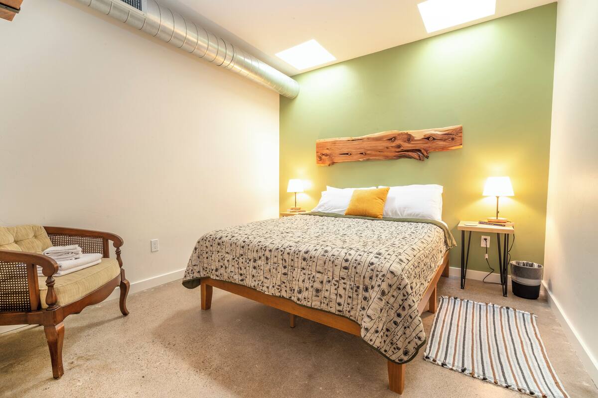 Quarto com cama, luminárias de cabeceira, painel de cama de madeira, um banquinho e um tapete pequeno.