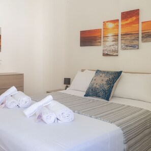 Uma cama bem arrumada com toalhas dobradas, cabeceira com almofada decorativa e três quadros de pôr do sol na parede.