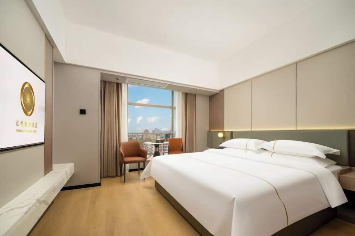 Quarto de hotel com cama, escrivaninha, cadeira, janela com cortinas e vista para a cidade.