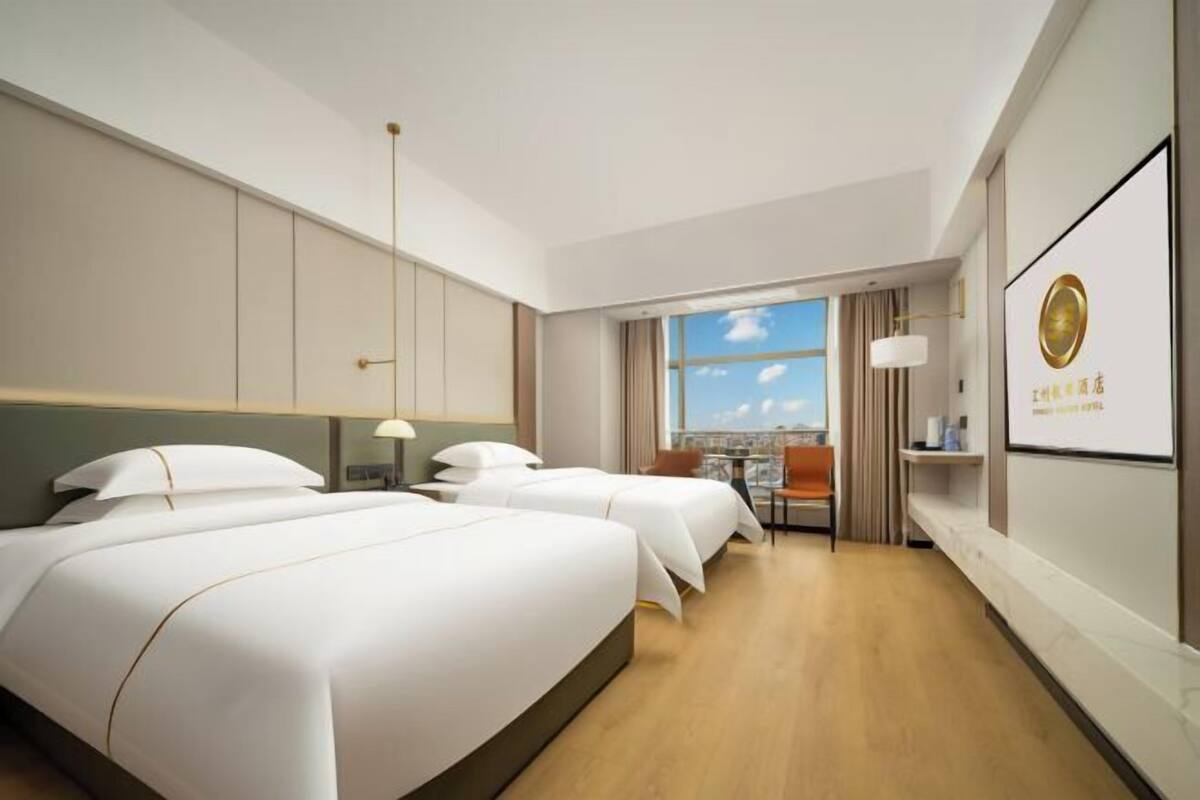 Quarto de hotel com duas camas, uma escrivaninha, uma cadeira, janela com vista para a cidade e tela fixada na parede.