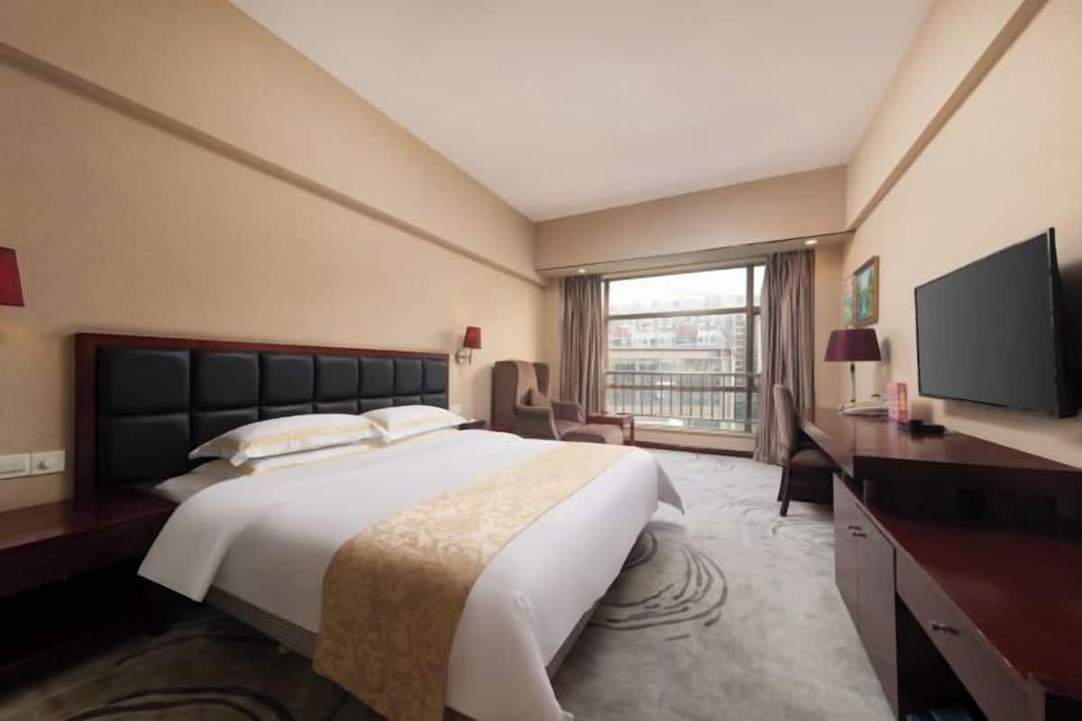 Quarto de hotel com uma cama grande, uma escrivaninha, uma cadeira, uma televisão e uma janela com cortinas.